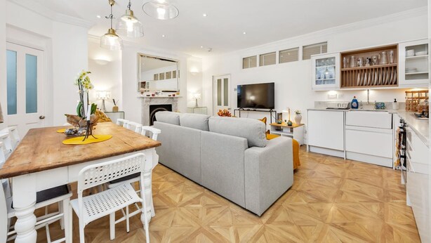 Uma sala de estar moderna com uma mesa de jantar de madeira, cadeiras brancas, um sofá cinza e uma TV.