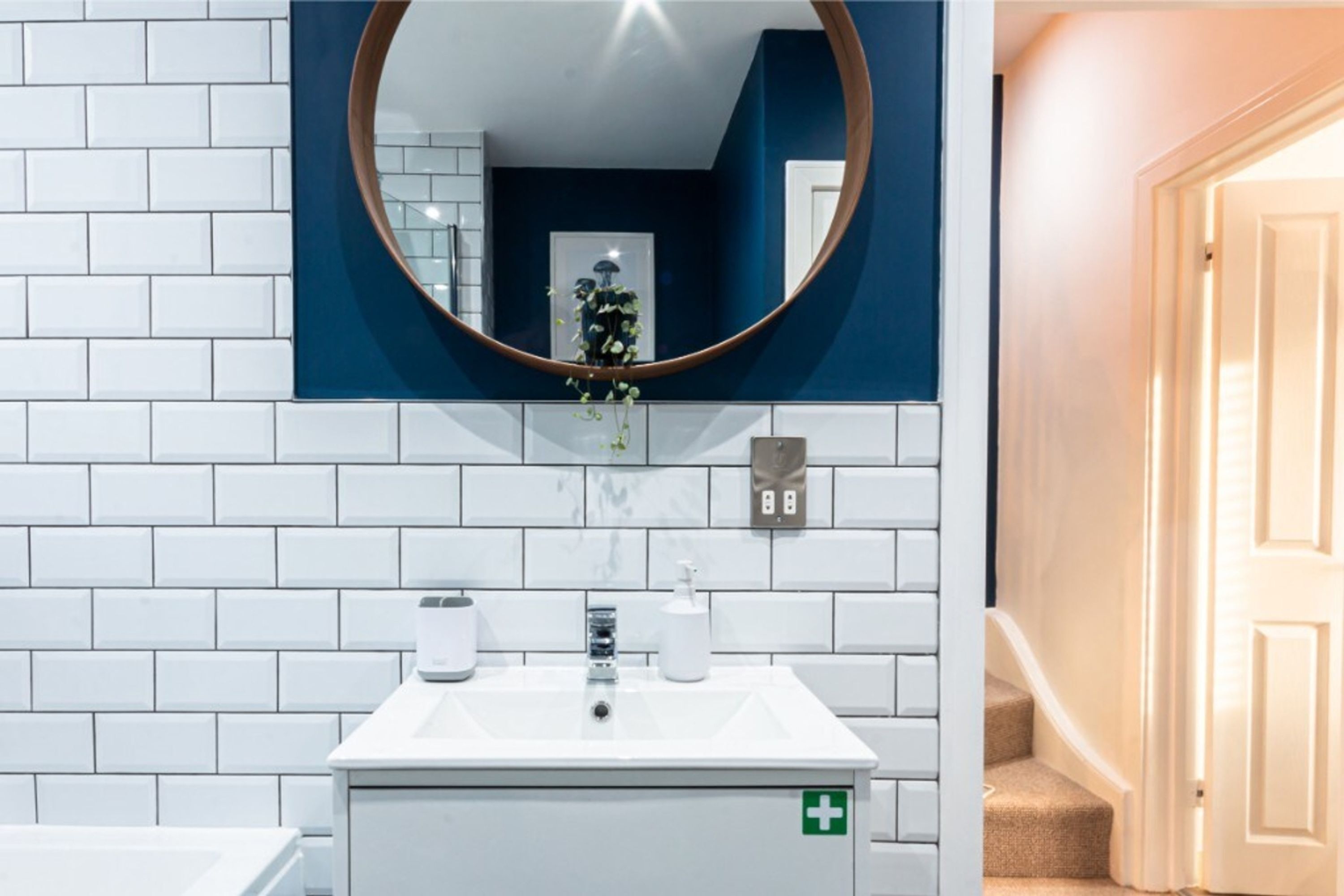 Casa de banho com paredes revestidas de azulejos brancos, uma pia branca, um espelho com moldura de madeira e um sinal de saída de emergência verde.