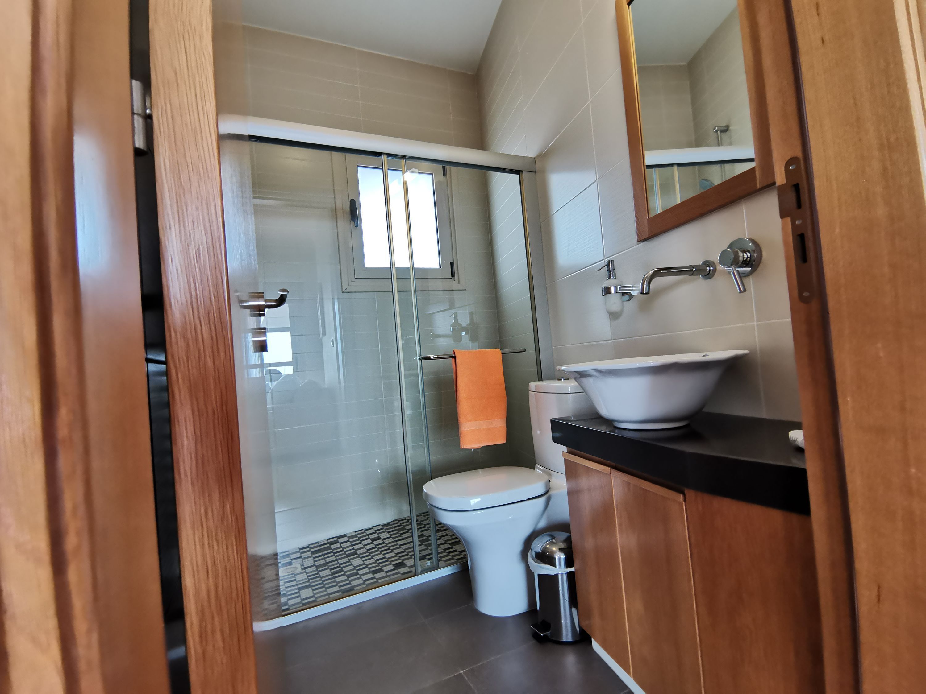 Casa de banho moderna com cabine de duche em vidro, sanita branca e toucador de madeira com lavatório branco.
