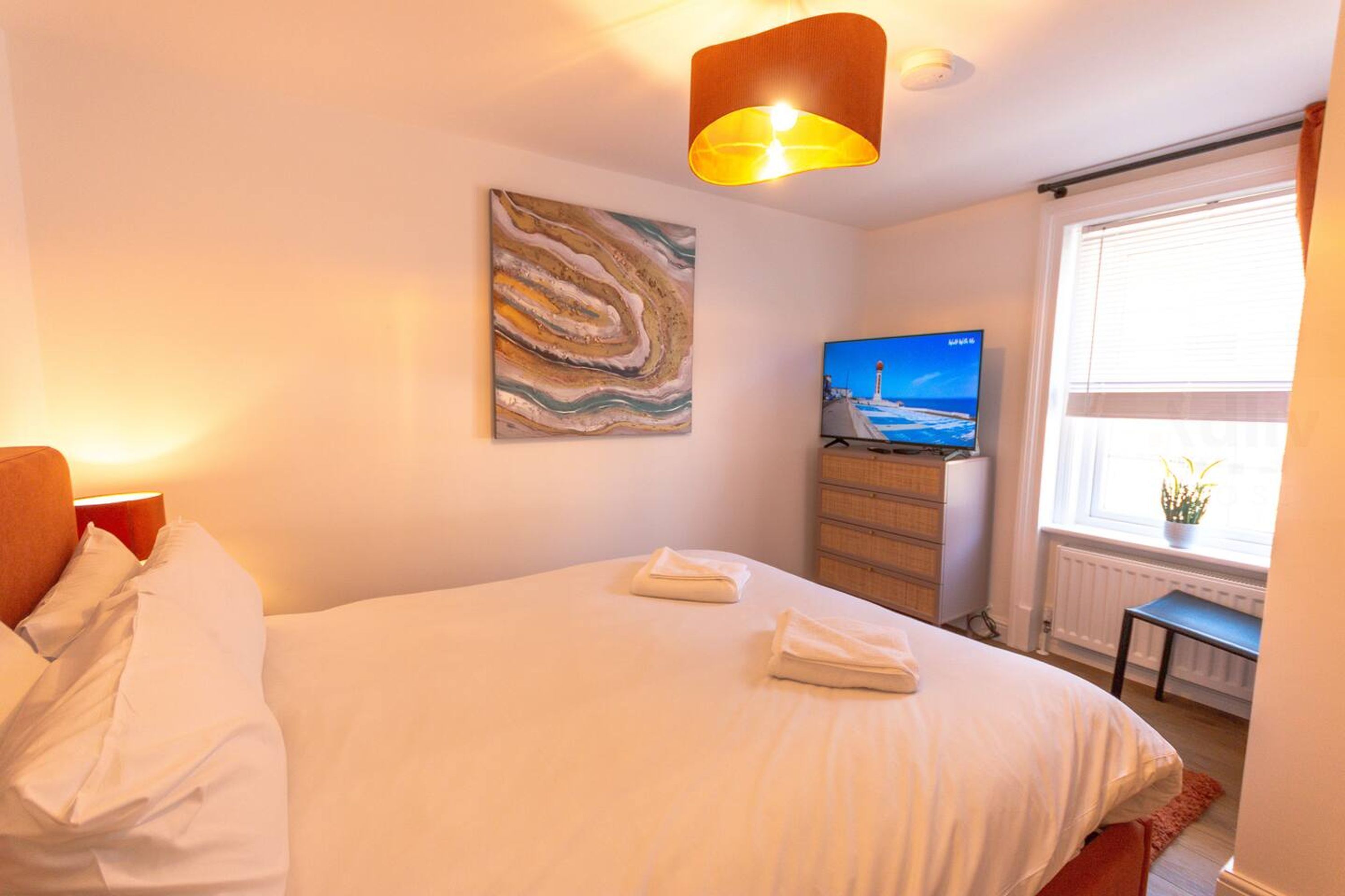 Quarto de hotel com cama, televisão, quadro e janela com persianas.