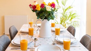 Mesa de café da manhã posta com pratos, copos de suco de laranja, jarra de suco de laranja e um vaso com flores coloridas.