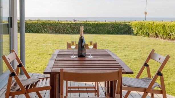 Uma mesa de madeira com duas cadeiras, uma garrafa de vinho e dois copos numa varanda com vista para o mar.