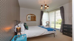 Quarto com cama, luminária de cabeceira, cômoda e janela com cortinas.