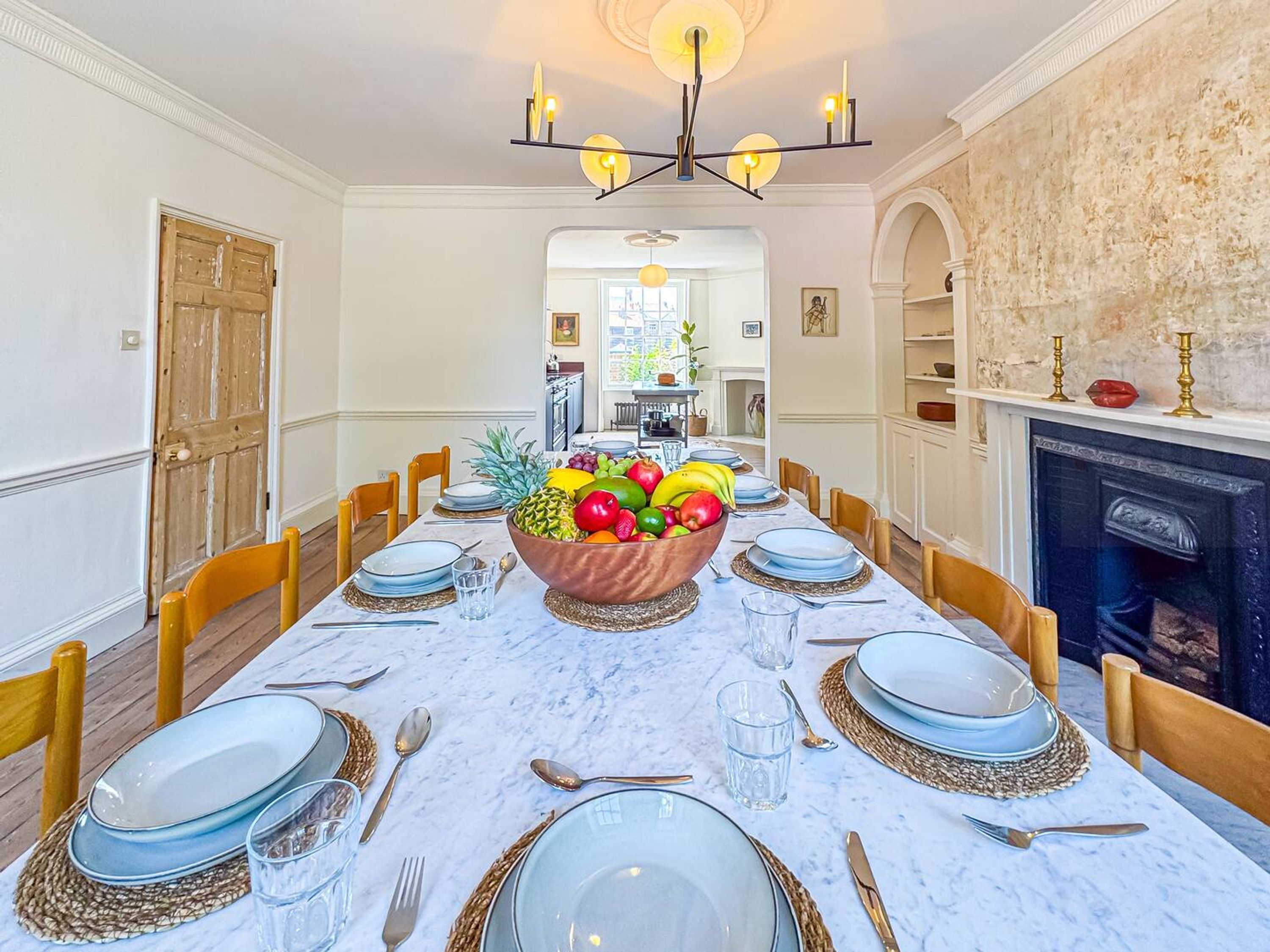 Uma sala de jantar com uma mesa comprida posta para refeição, um centro de mesa com frutas e uma lareira.