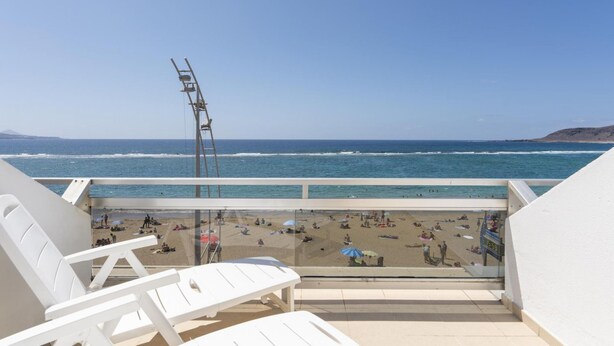 Varanda com cadeiras brancas com vista para uma praia com pessoas e guarda-sóis.