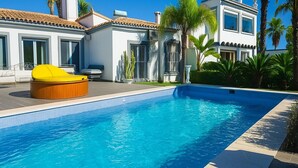 Piscina com hidromassagem, palmeiras e um edifício branco com telhado em azulejos.