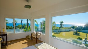 Quarto com uma cama grande, uma cadeira, uma mesinha e vista para um jardim bem cuidado e para o oceano.