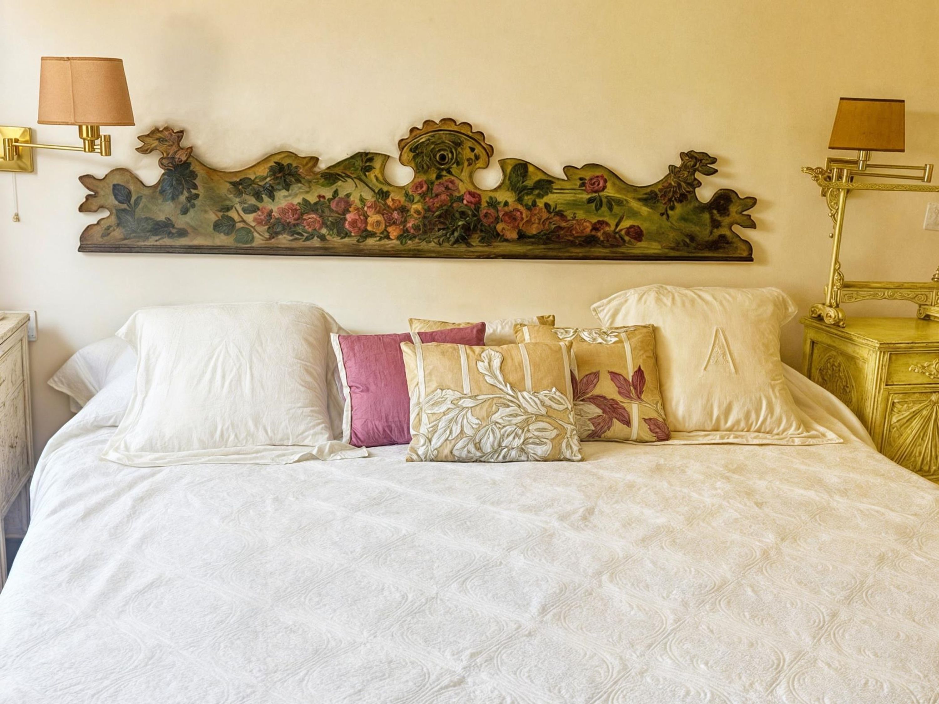 Cama bem arrumada com lençóis brancos e almofadas decorativas, um painel floral na parede acima e uma mesa de cabeceira dourada com abajur.