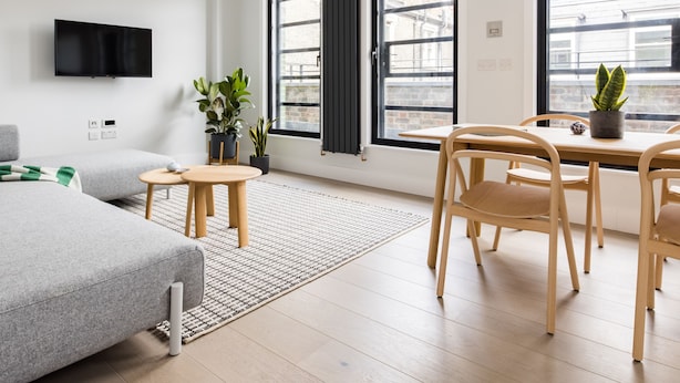 Uma sala de estar moderna com sofá, mesa de jantar com cadeiras, TV montada na parede e grandes janelas com vista para edifícios.