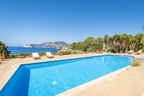 Uma piscina retangular com água azul cristalina, ladeada por azulejos bege. Existem espreguiçadeiras e plantas em vasos ao redor da piscina. Ao fundo, vê-se o mar e colinas distantes.