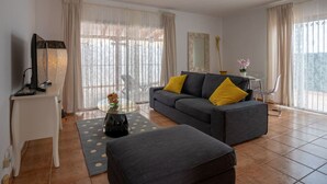 Uma sala de estar com um sofá cinzento, uma pequena mesa lateral, uma mesa de centro de vidro e uma janela grande com cortinas leves.