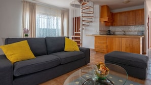 Uma sala de estar moderna com um sofá cinzento, uma mesa de centro de vidro e um travesseiro amarelo. Uma escada em espiral leva a uma cozinha de madeira com armários e bancada.