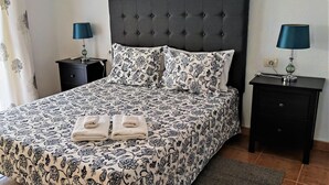Cama bem arrumada com colcha estampada, dois travesseiros iguais e uma toalha dobrada. Há duas mesas de cabeceira, cada uma com uma luminária e uma tomada.