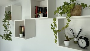 Uma parede branca com três prateleiras brancas, cada uma com livros, uma planta em vaso e um pequeno relógio.