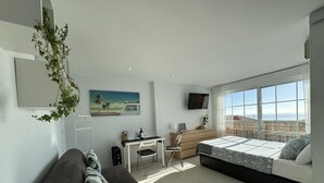 Quarto moderno com cama, escrivaninha e sofá. Janela com vista para a praia.