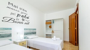 Quarto de hotel com duas camas, um guarda-roupa de madeira, uma prateleira com toalhas e uma parede com texto decorativo.