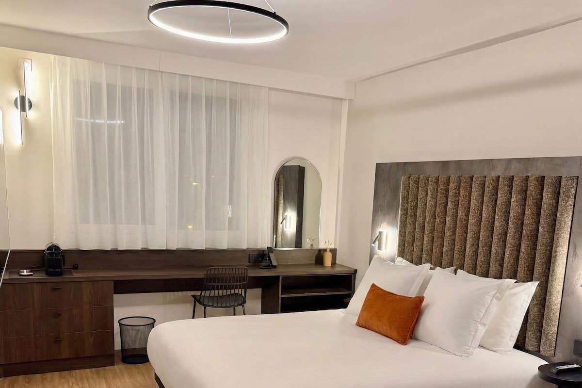 Quarto de hotel moderno com uma cama grande, uma escrivaninha com cadeira, um pequeno cesto de lixo e um espelho amplo.