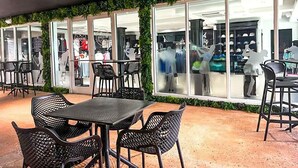 Uma área externa com mesas e cadeiras, uma loja com araras de roupas e uma treliça verde na parede.