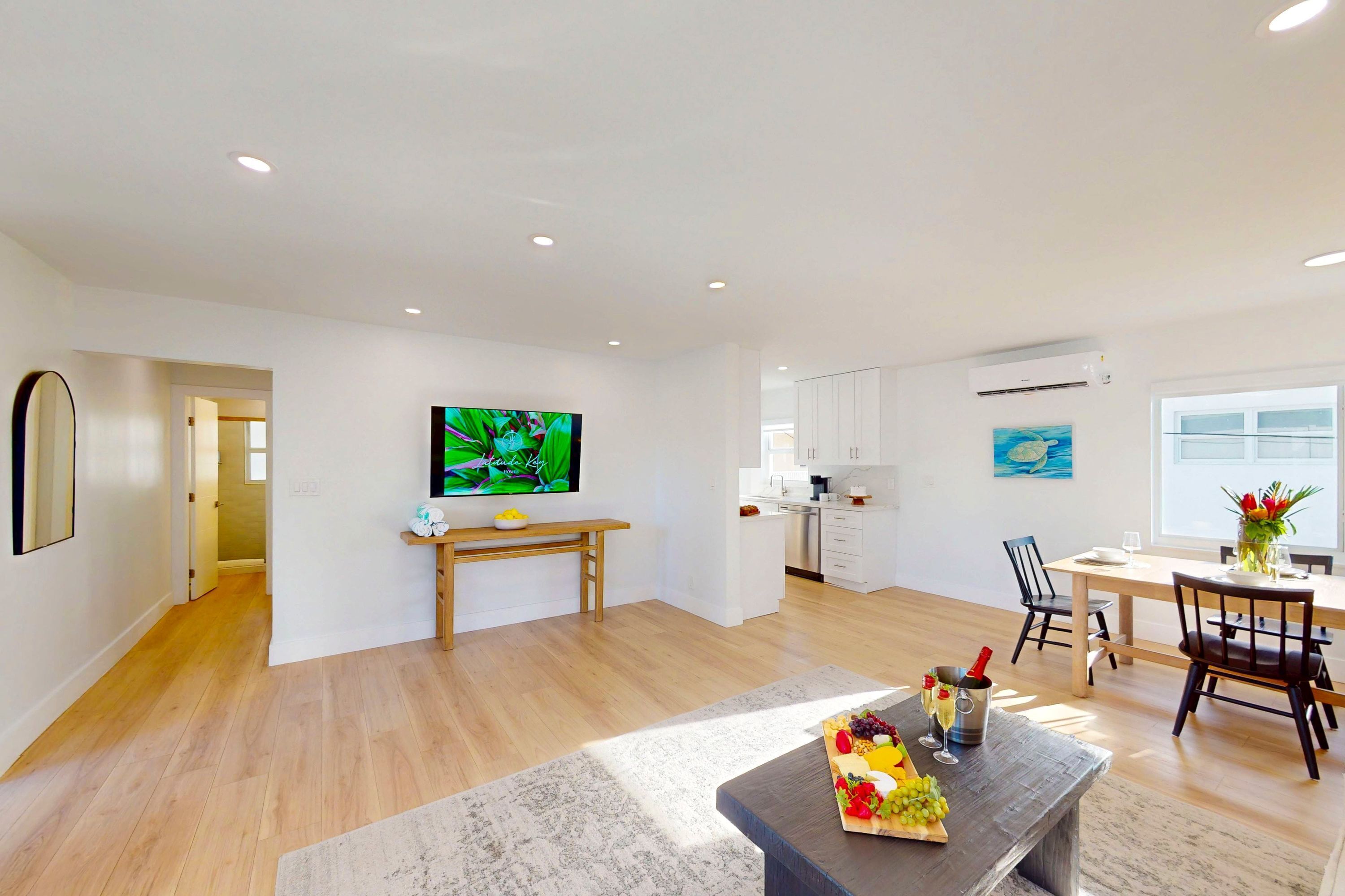 Uma sala de estar moderna com piso de madeira, área de jantar com mesa e cadeiras, cozinha ao fundo e uma TV em um suporte.
