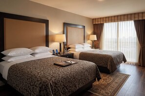 Quarto de hotel com duas camas, uma janela ampla com cortinas, mesa de cabeceira com luminárias e um tapete.