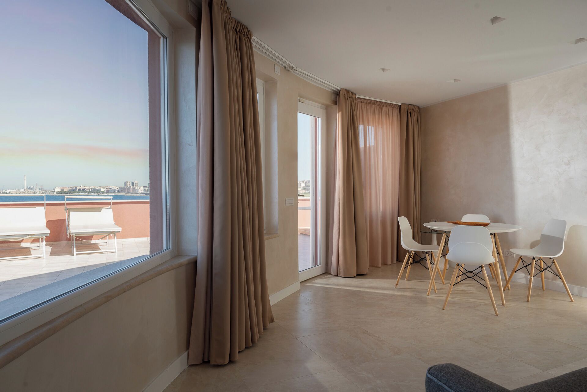 Quarto de hotel moderno com varanda, área de refeições com cadeiras e mesa brancas, e vista para a cidade.