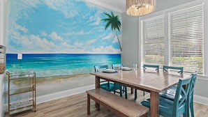 Uma área de refeições com um mural de praia, mesa de madeira, cadeiras verde-água e luminária pendente.