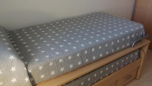 Quarto pequeno com uma cama de solteiro, colcha estampada com estrelas e mesa de cabeceira de madeira com gaveta.