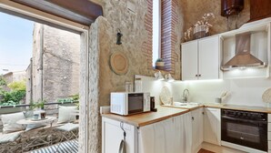 Cozinha compacta com armários brancos, forno embutido e micro-ondas. Há uma pequena área de refeições com mesa e cadeiras ao ar livre.
