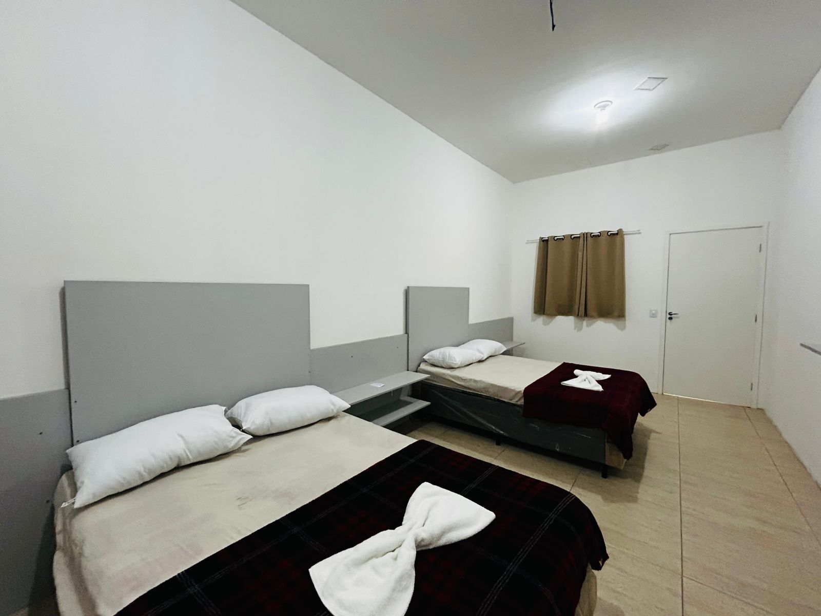 Quarto com duas camas, cada uma com cabeceira, uma janela com cortinas e uma porta.