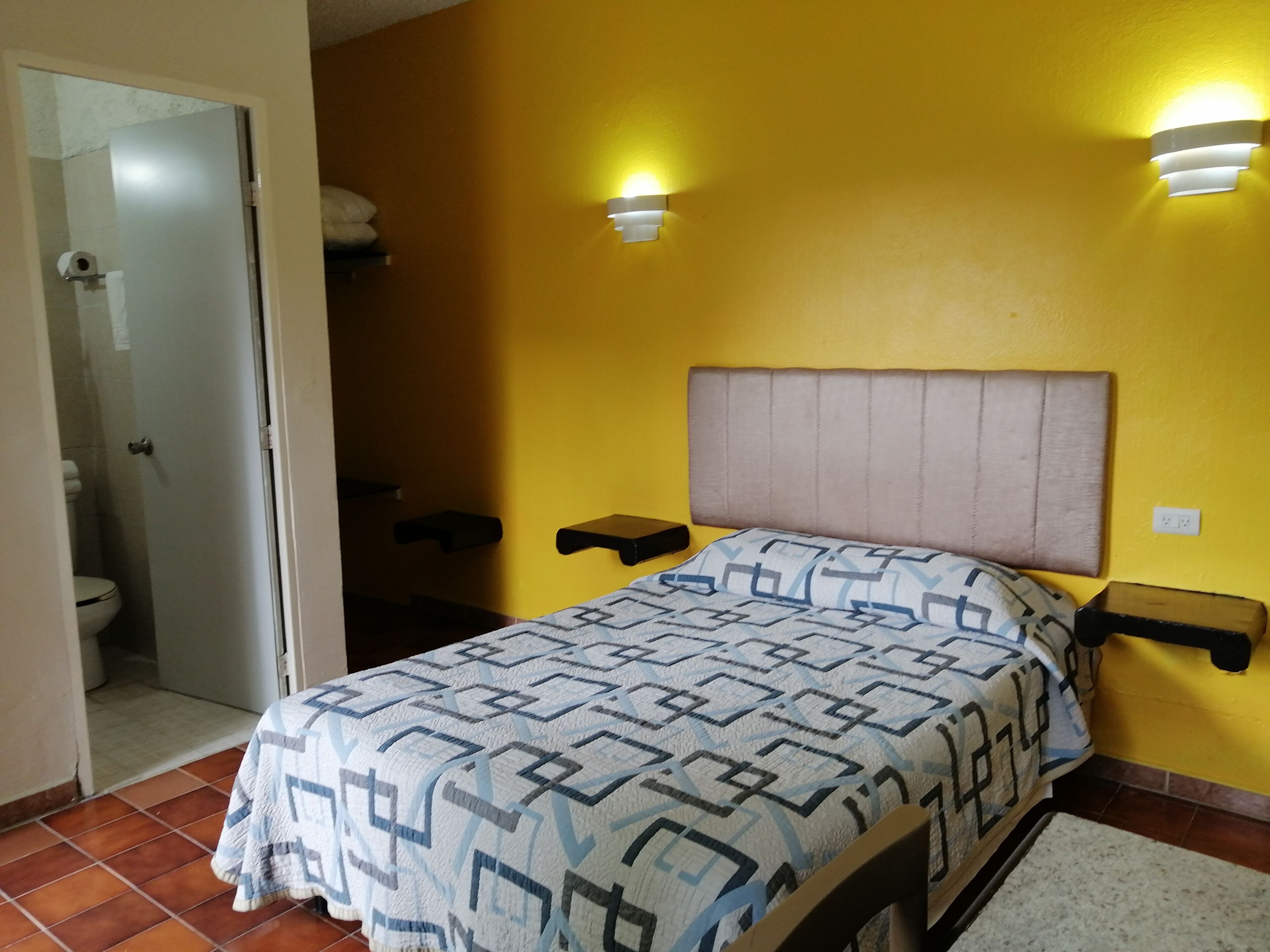 Quarto com cama, mesa de cabeceira, luminária de parede e painel de cabeceira.