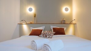 Cama bem arrumada com toalhas enroladas e duas almofadas, mesa de cabeceira com um vaso e abajur, e luminárias circulares fixadas na parede.