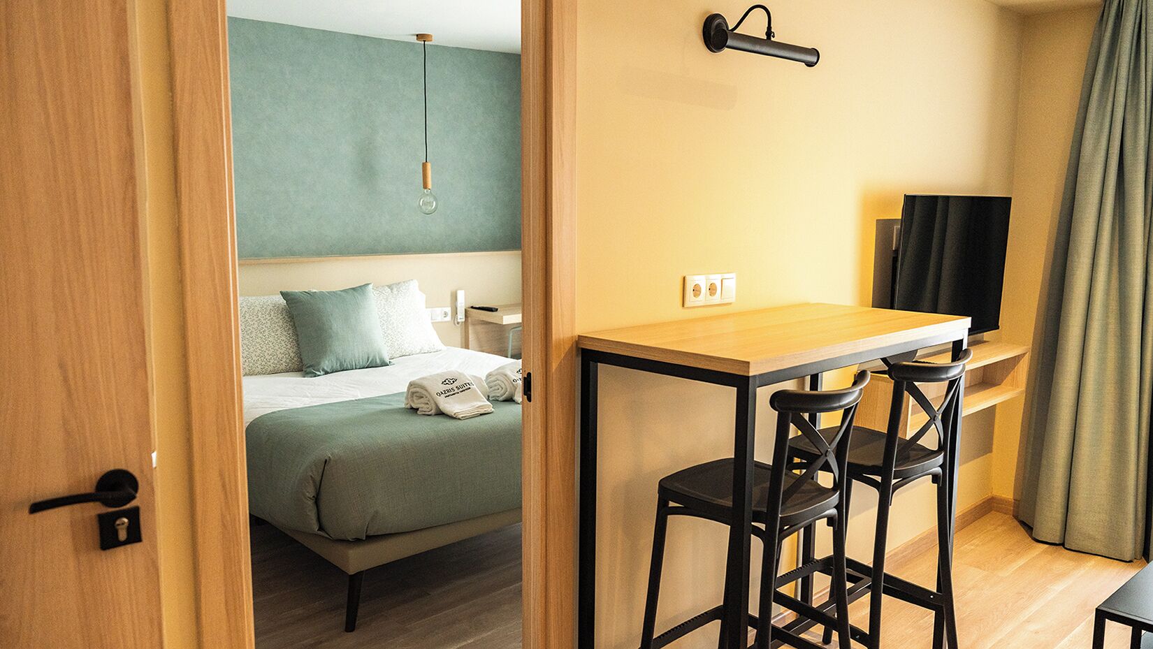 Quarto de hotel com uma cama, uma mesa com duas cadeiras, uma televisão e uma porta.