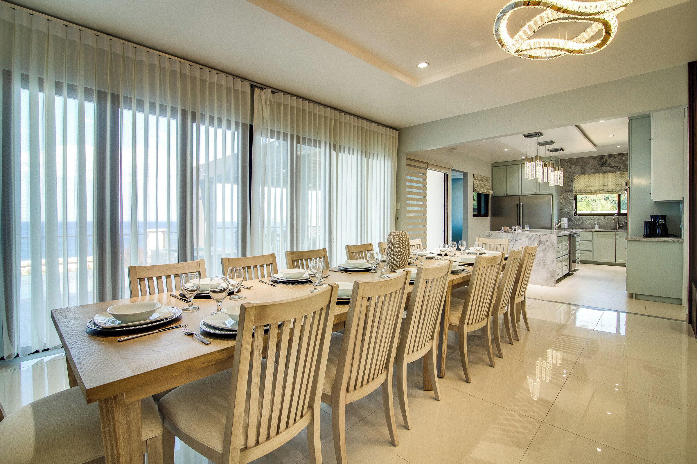 Uma sala de jantar com mesa de madeira posta para seis, janelas amplas com cortinas leves e uma cozinha moderna ao fundo.