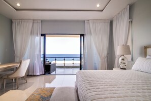 Quarto com uma cama grande, uma escrivaninha, uma cadeira, uma luminária e vista para o oceano.