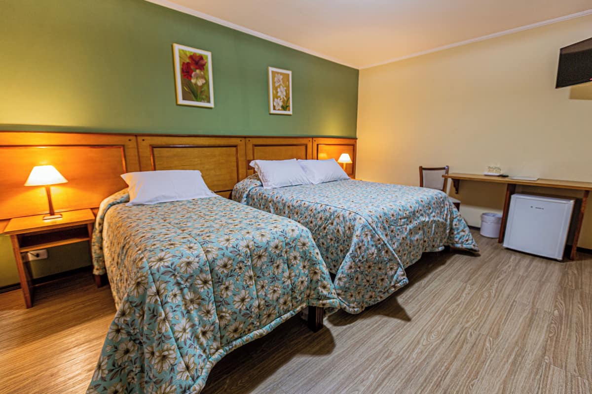Quarto de hotel com duas camas, uma escrivaninha, uma cadeira, televisão e um cesto de lixo.