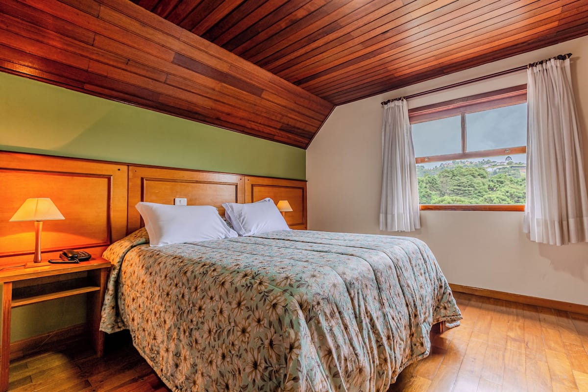 Quarto de hotel com teto de madeira, cama com roupa de cama floral, mesa de cabeceira com abajur e janela com cortinas.