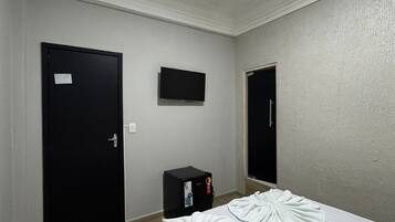 Quarto de hotel com duas portas, uma televisão fixada na parede e uma cama com duas toalhas brancas.