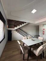 Um interior moderno com área de refeições, mesa branca e quatro cadeiras, escada com corrimão preto e um extintor de incêndio fixado na parede.