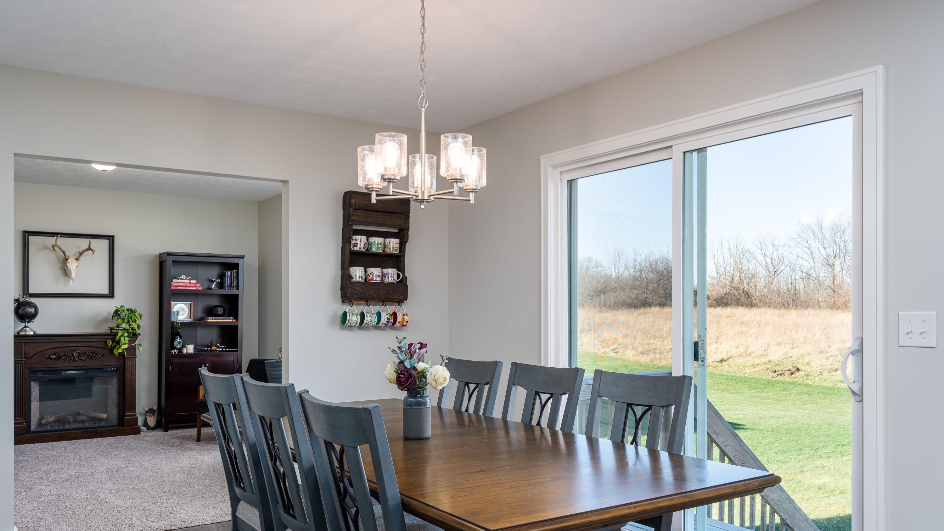 Uma sala de jantar com mesa de madeira, seis cadeiras, um candelabro, lareira, estante de livros e uma grande janela com vista para o exterior.