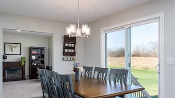 Uma sala de jantar com mesa de madeira, seis cadeiras, um candelabro, lareira, estante de livros e uma grande janela com vista para o exterior.