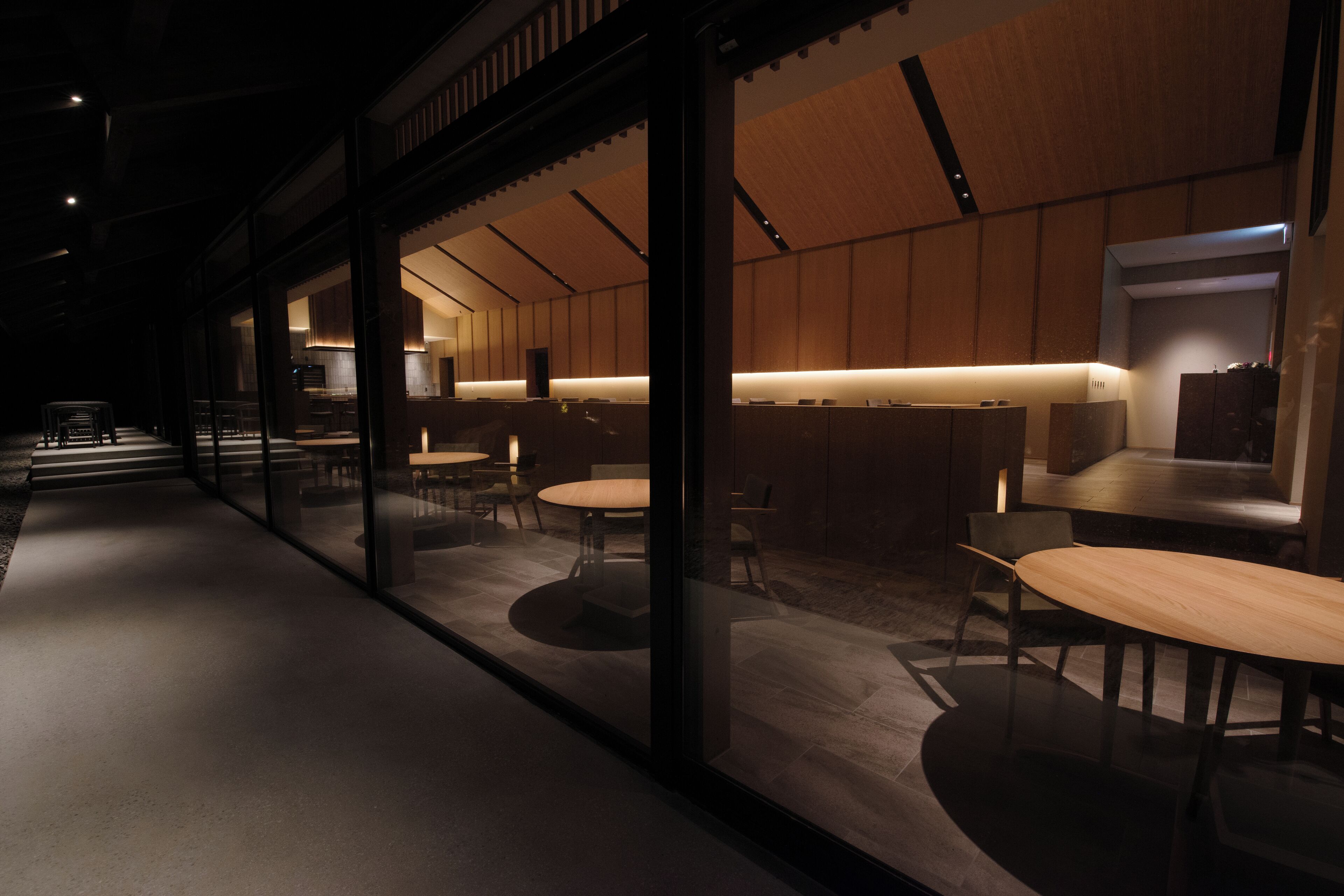 Um interior moderno com divisórias de vidro, piso de madeira e uma área de estar central com mesas e cadeiras.