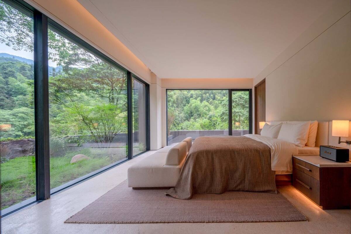 Quarto moderno com uma janela ampla, cama, mesa de cabeceira e vista para um jardim exuberante.