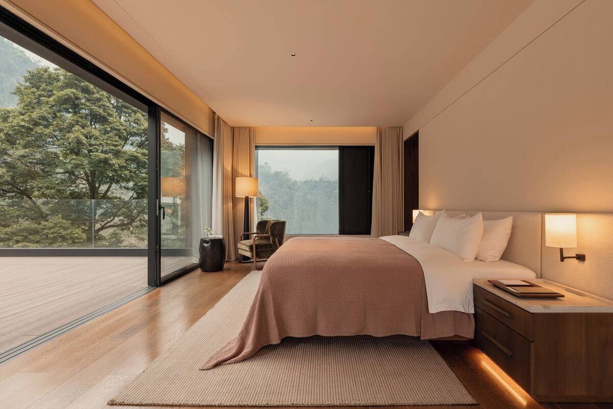 Quarto moderno com uma grande janela, uma cama, mesas de cabeceira e vista para árvores.