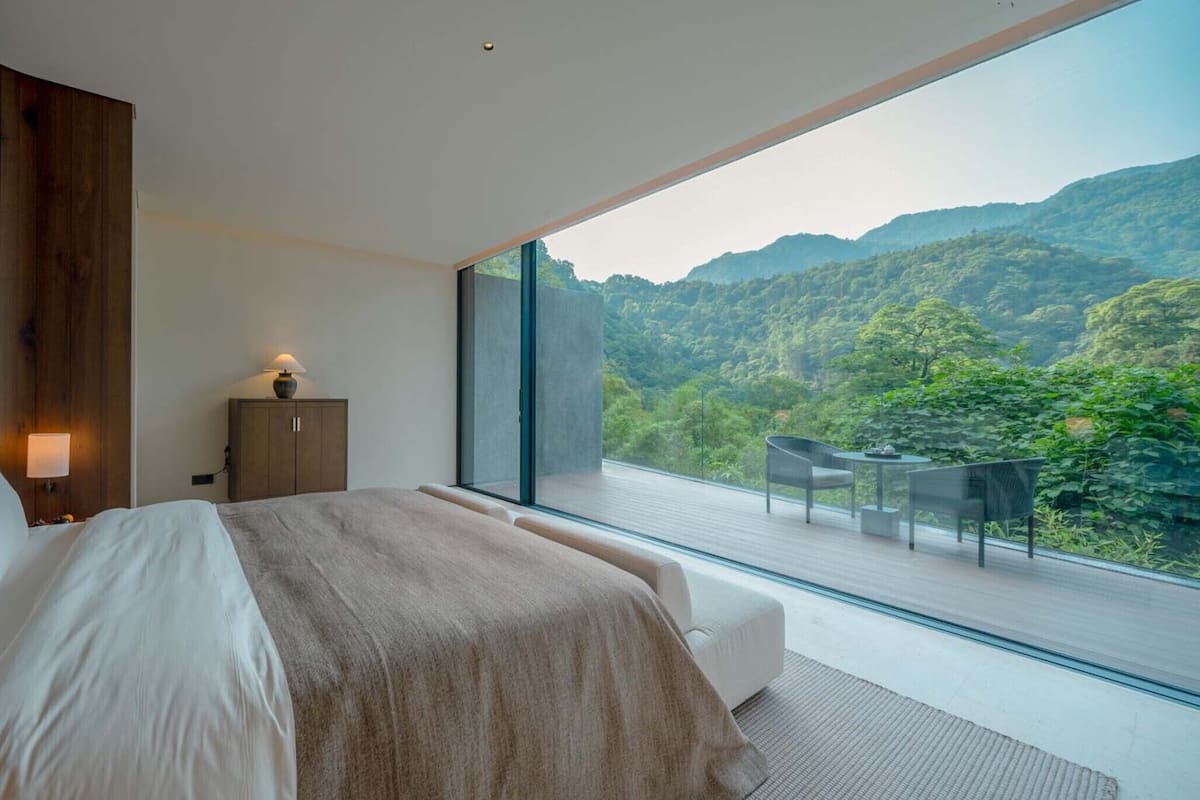 Quarto com uma janela ampla, uma cama, mesa de cabeceira e uma luminária, além de vista para uma encosta arborizada.