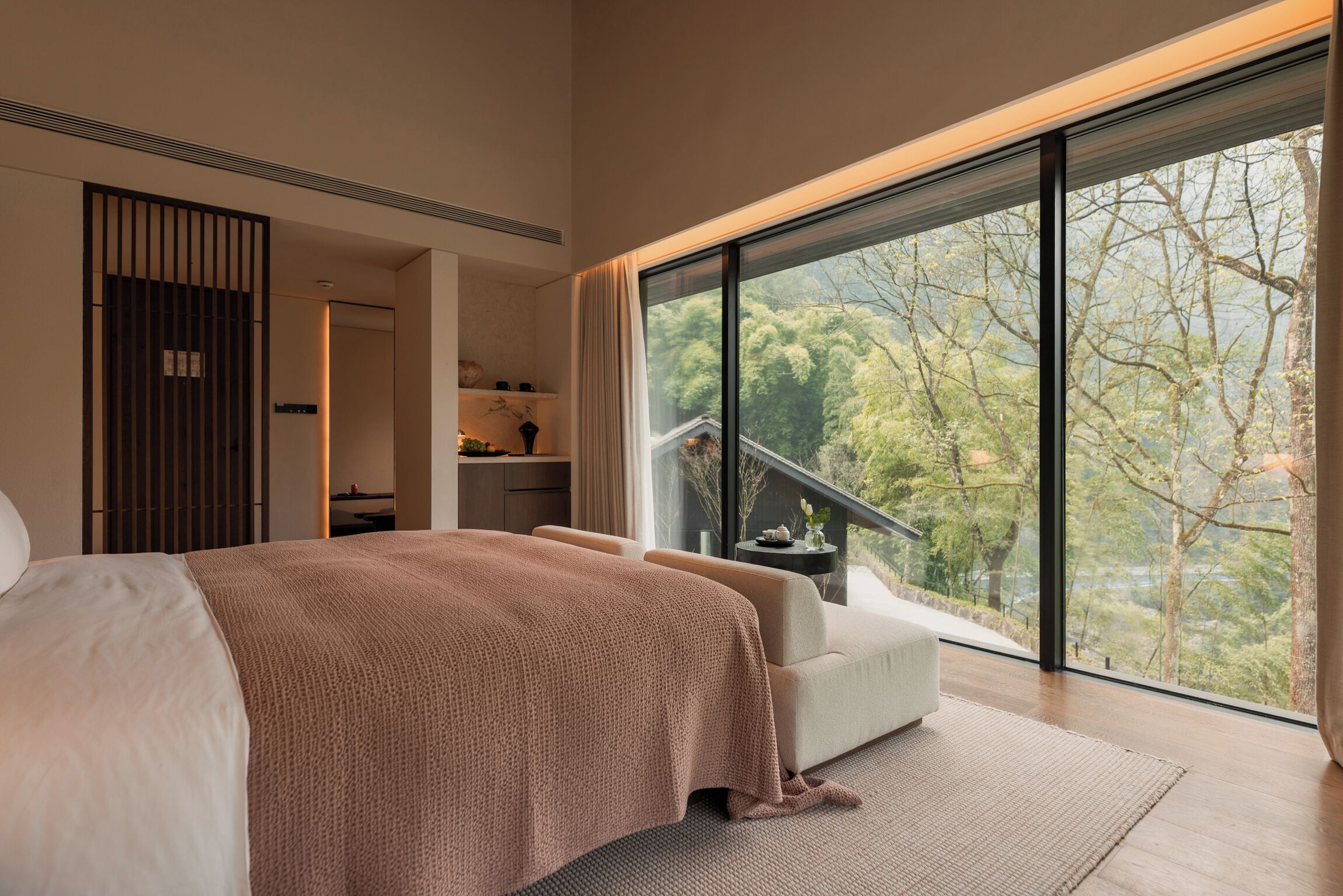 Quarto moderno com uma cama grande, um sofá, uma mesinha e vista para árvores através de janelas amplas.