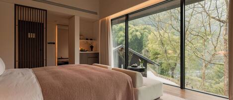 Quarto moderno com uma cama grande, um sofá, uma mesinha e vista para árvores através de janelas amplas.
