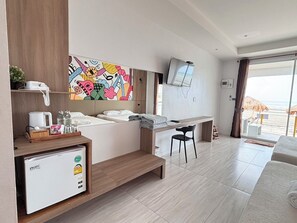 Quarto de hotel moderno com cozinha compacta, uma grande janela com vista para a praia, um quadro abstrato colorido e uma TV de tela plana fixada na parede.