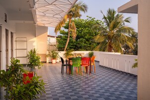 Terraço com piso xadrez, mesa com cadeiras, plantas em vasos e palmeiras ao fundo.