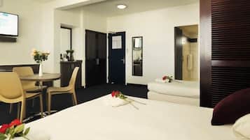 Quarto de hotel com cama, escrivaninha e cadeira. Há um relógio na parede e uma televisão montada sobre a escrivaninha.