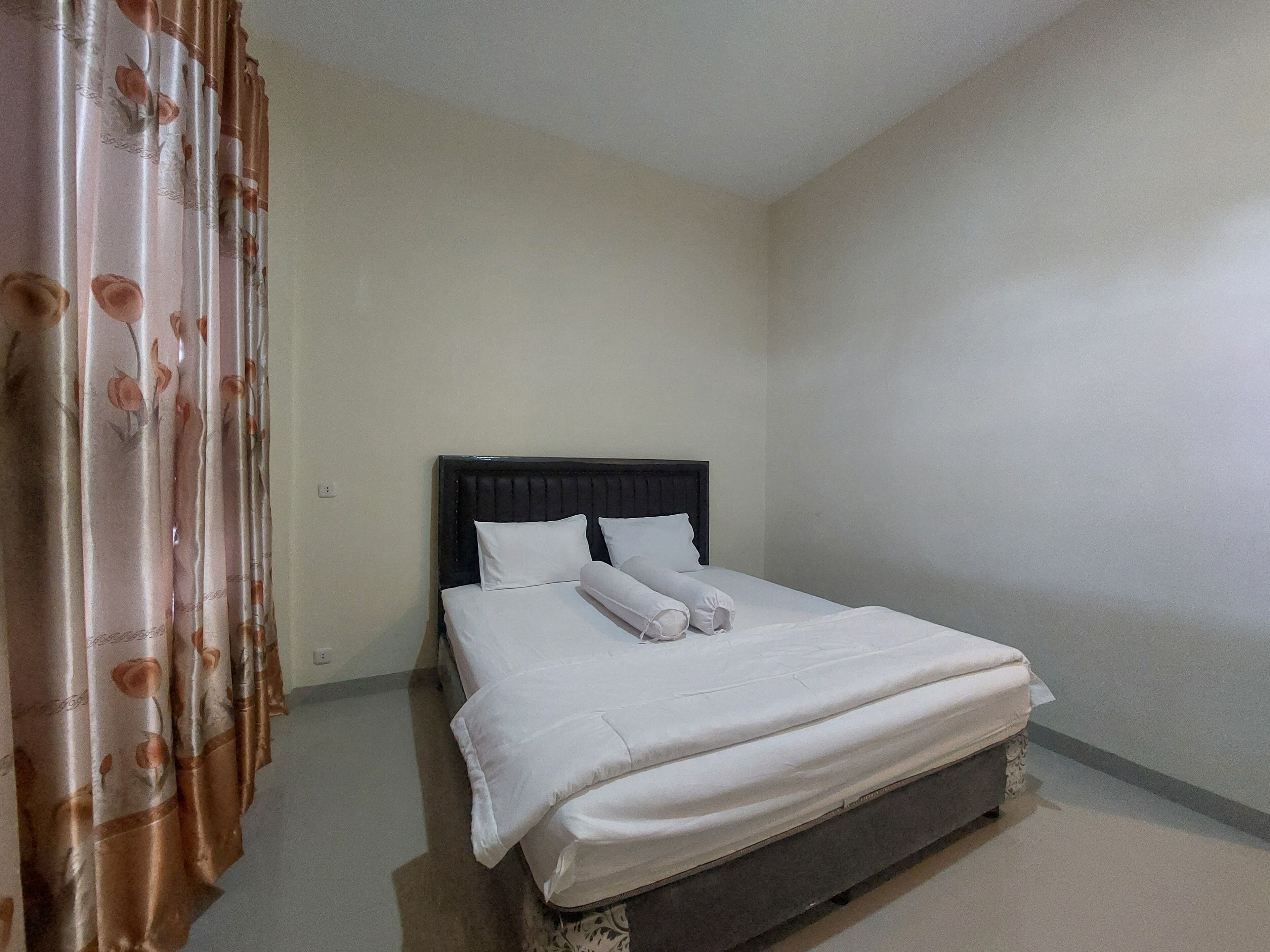 Cama bem arrumada com lençóis e almofadas brancos, cabeceira escura e cortina com padrão floral.